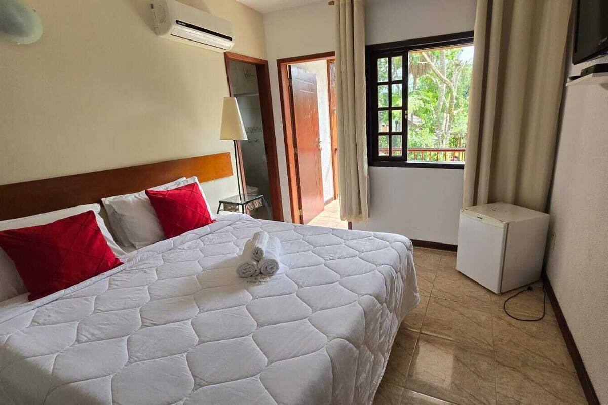 Quarto de hotel com cama, televisão, janela com vista para vegetação e aparelho de ar condicionado.