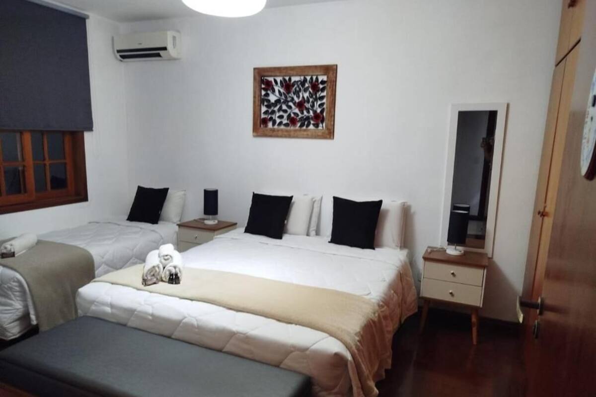 Quarto de hotel com uma cama grande, uma cama menor, um banco, uma mesa de cabeceira e um espelho, além de ar-condicionado fixado na parede.