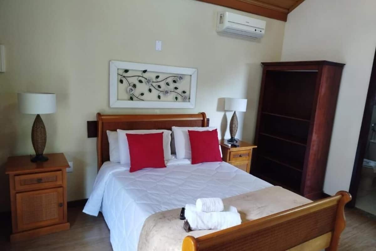 Uma cama bem arrumada com roupa de cama branca e vermelha, uma cabeceira de madeira e uma mesa de cabeceira com uma luminária.