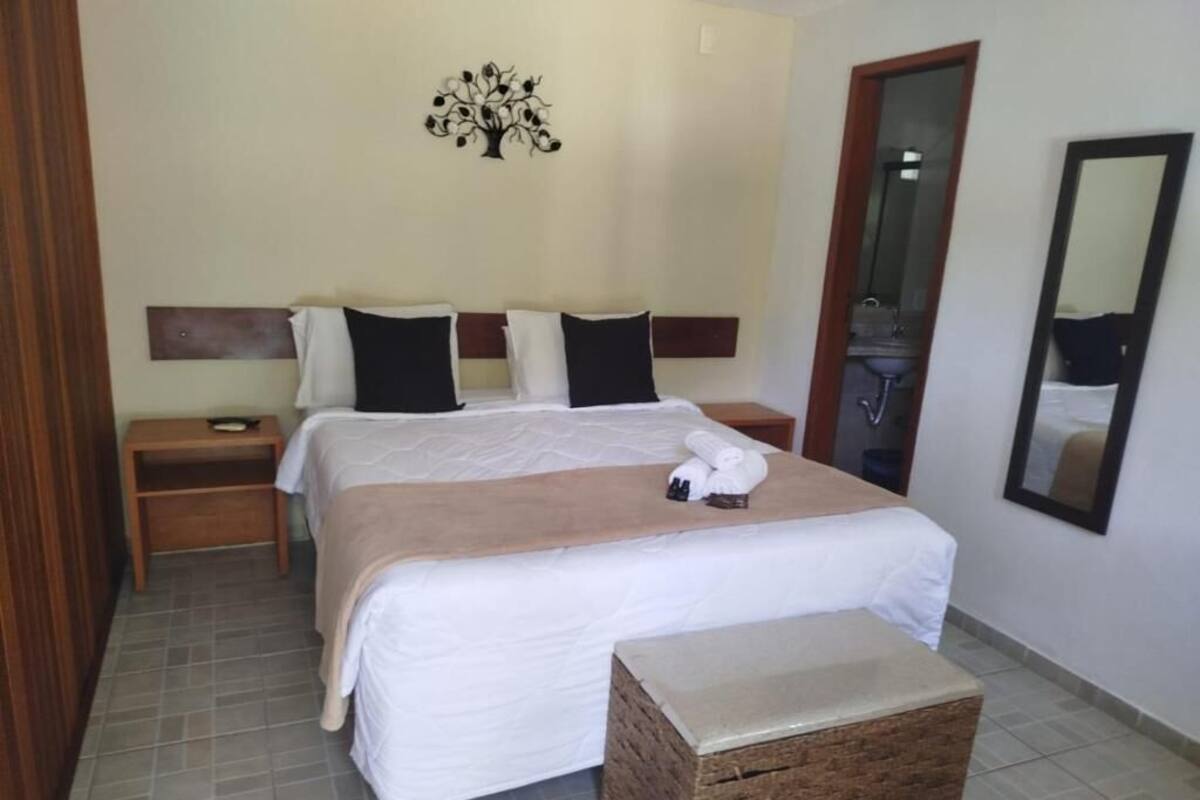 Uma cama bem arrumada com colcha branca e bege, um pequeno banco de madeira, uma mesinha de cabeceira, um espelho fixado na parede e um objeto decorativo na parede.