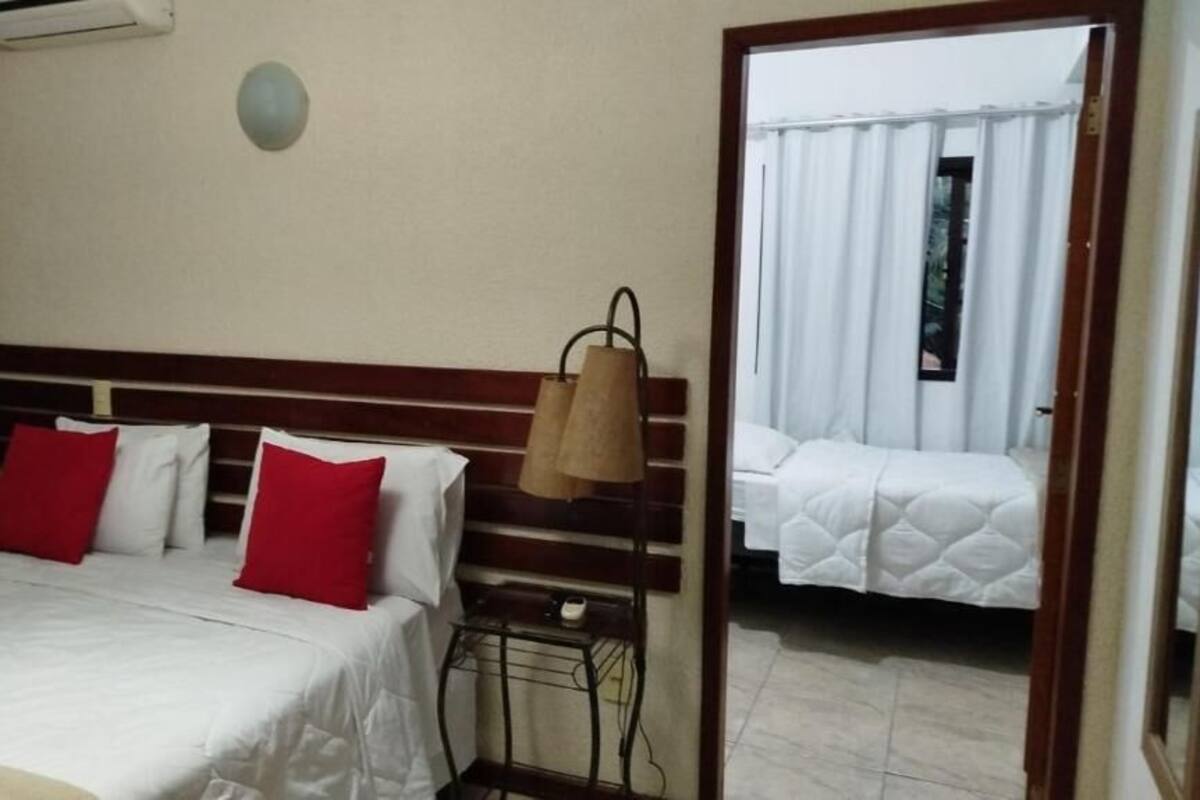 Quarto de hotel com cama, duas almofadas, mesa de cabeceira e espelho refletindo outro quarto.