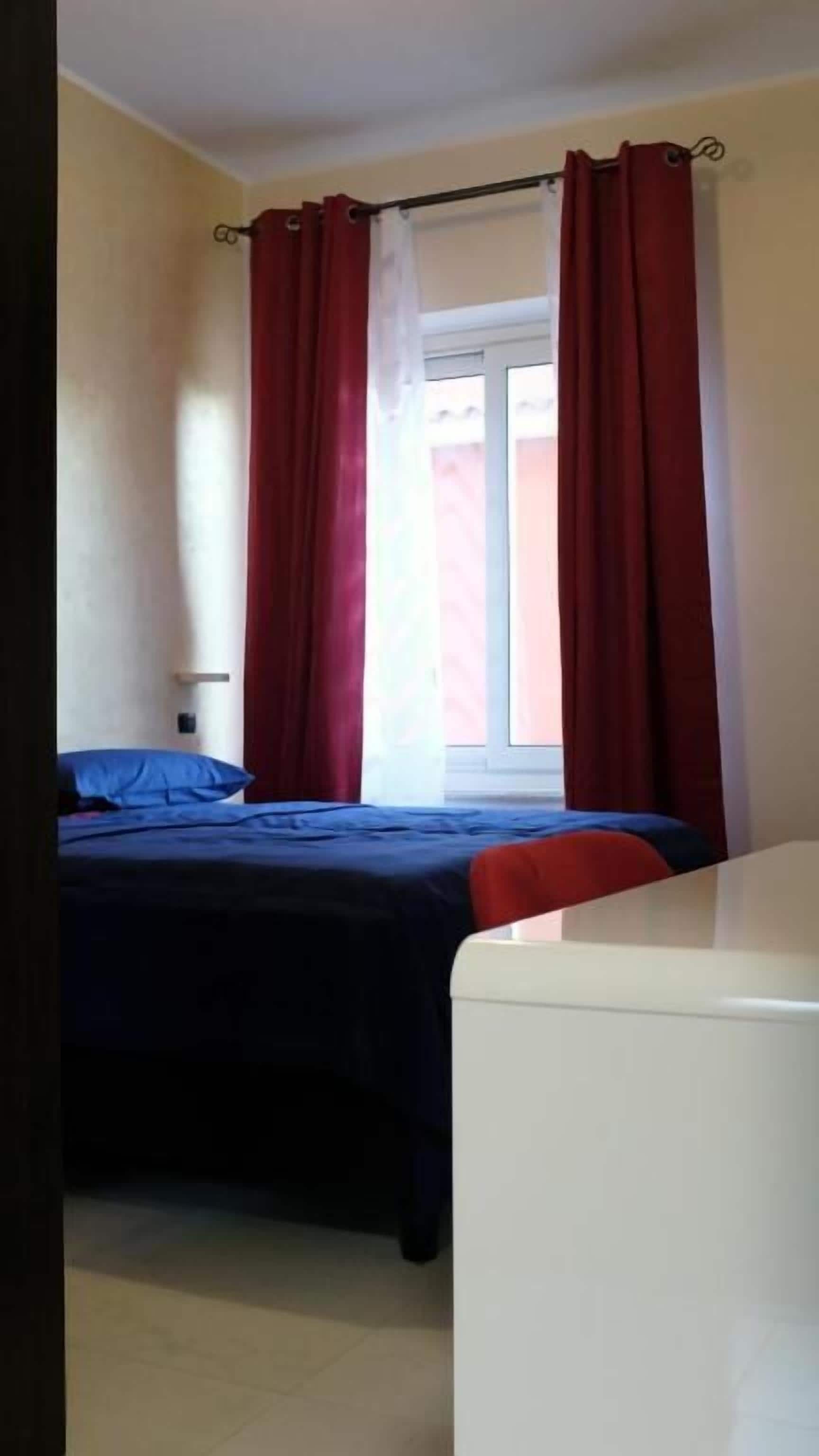 Quarto com uma cama, janela com cortinas vermelhas e mesa de cabeceira branca.