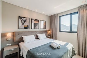 Quarto de hotel com cama, mesa de cabeceira, arte na parede e janela com cortinas.