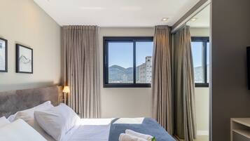 Quarto de hotel com cama, mesa de cabeceira, luminária, janela com cortinas e vista para a paisagem urbana.