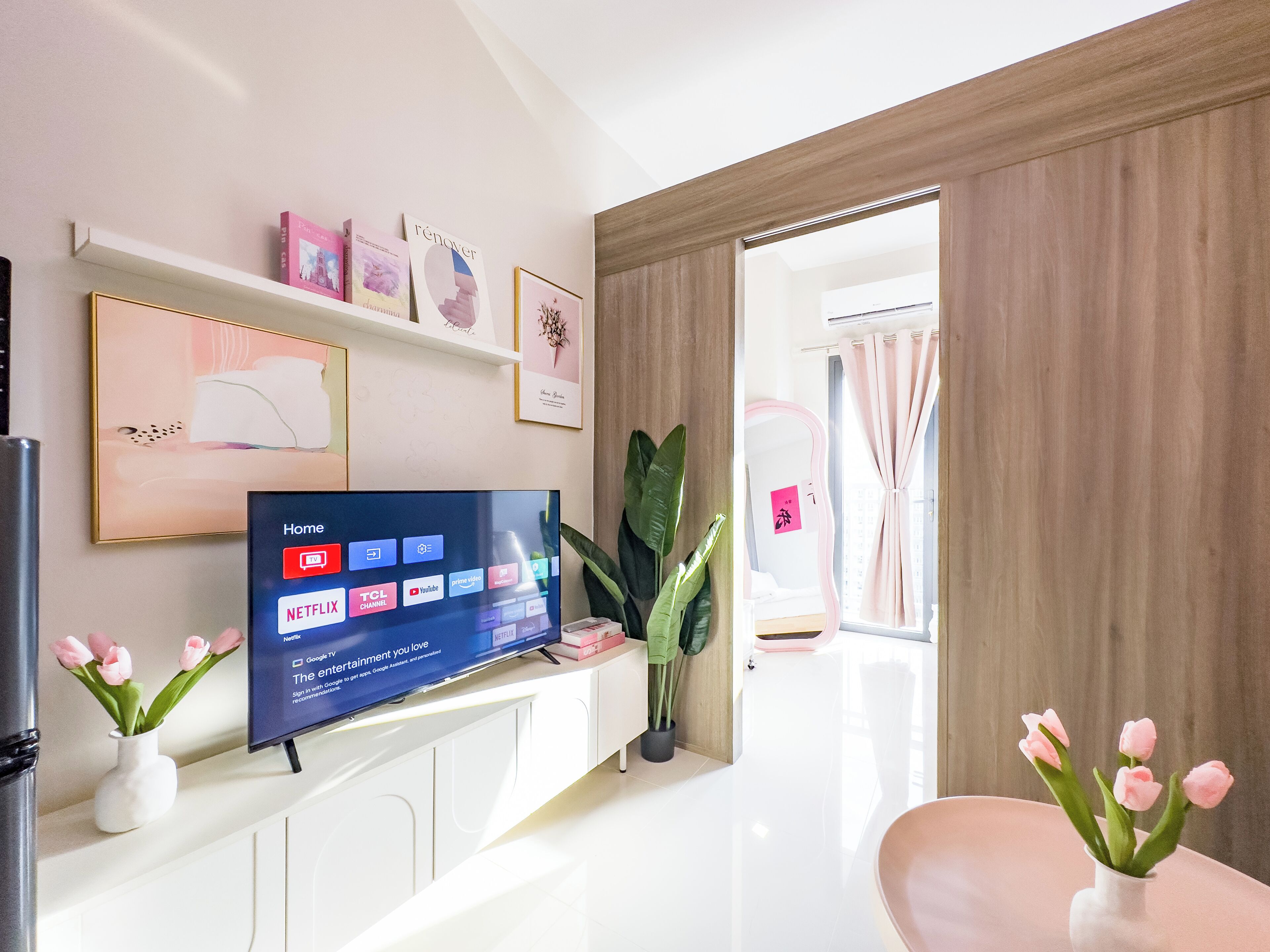 Uma sala de estar moderna com uma TV de tela plana exibindo serviços de streaming, um armário branco, uma planta em vaso e um vaso com flores rosa.