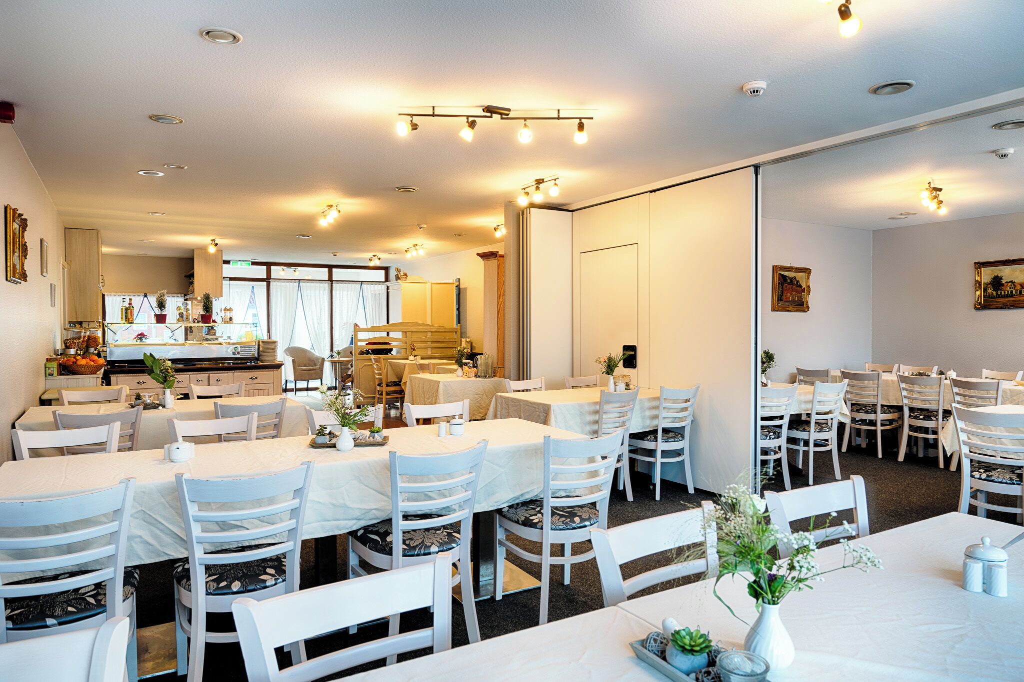 Uma sala de jantar com várias mesas e cadeiras, uma cozinha ao fundo e vista para o exterior através de grandes janelas.