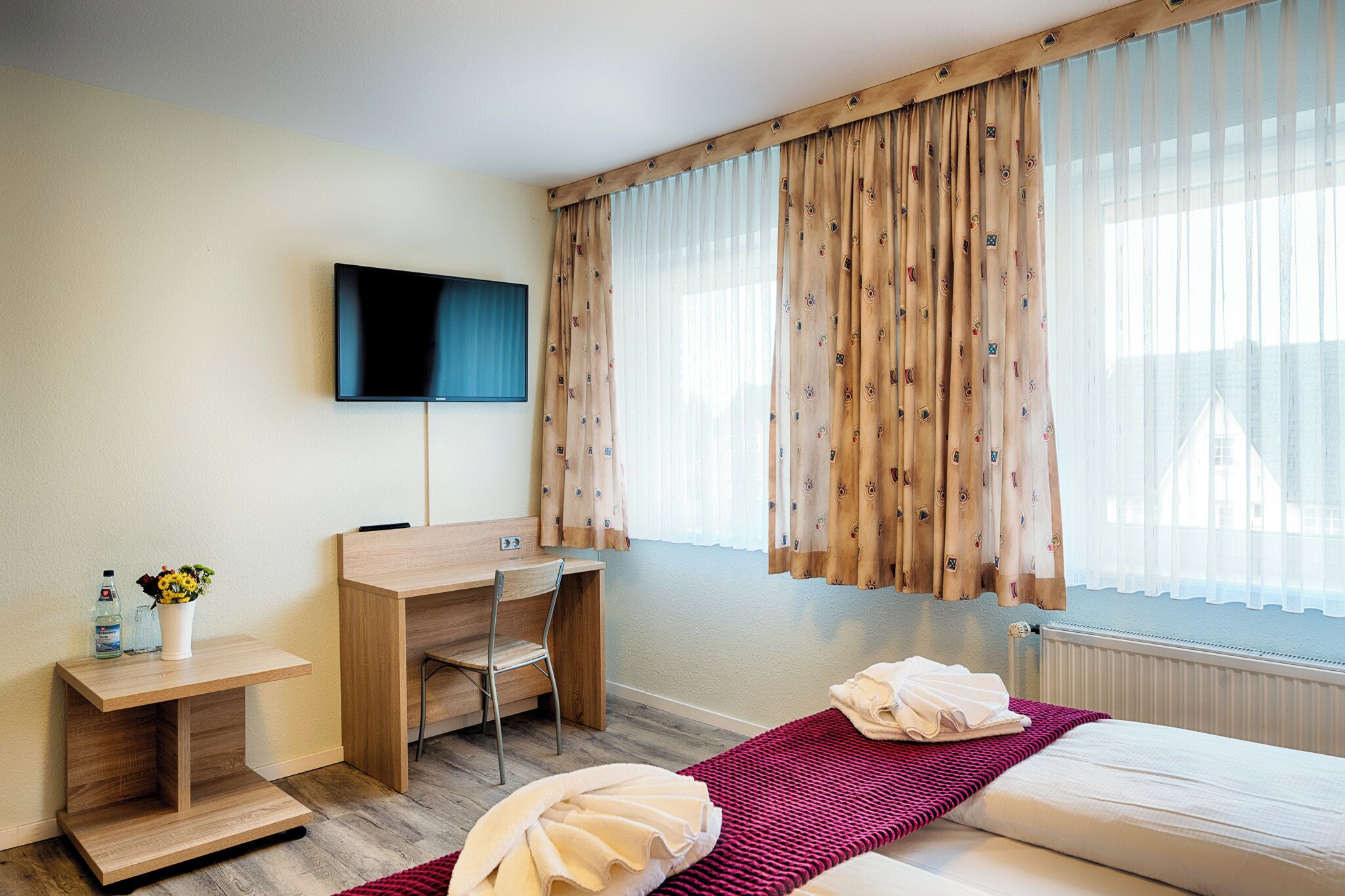 Quarto de hotel com cama, escrivaninha, cadeira, televisão e janela com cortinas.