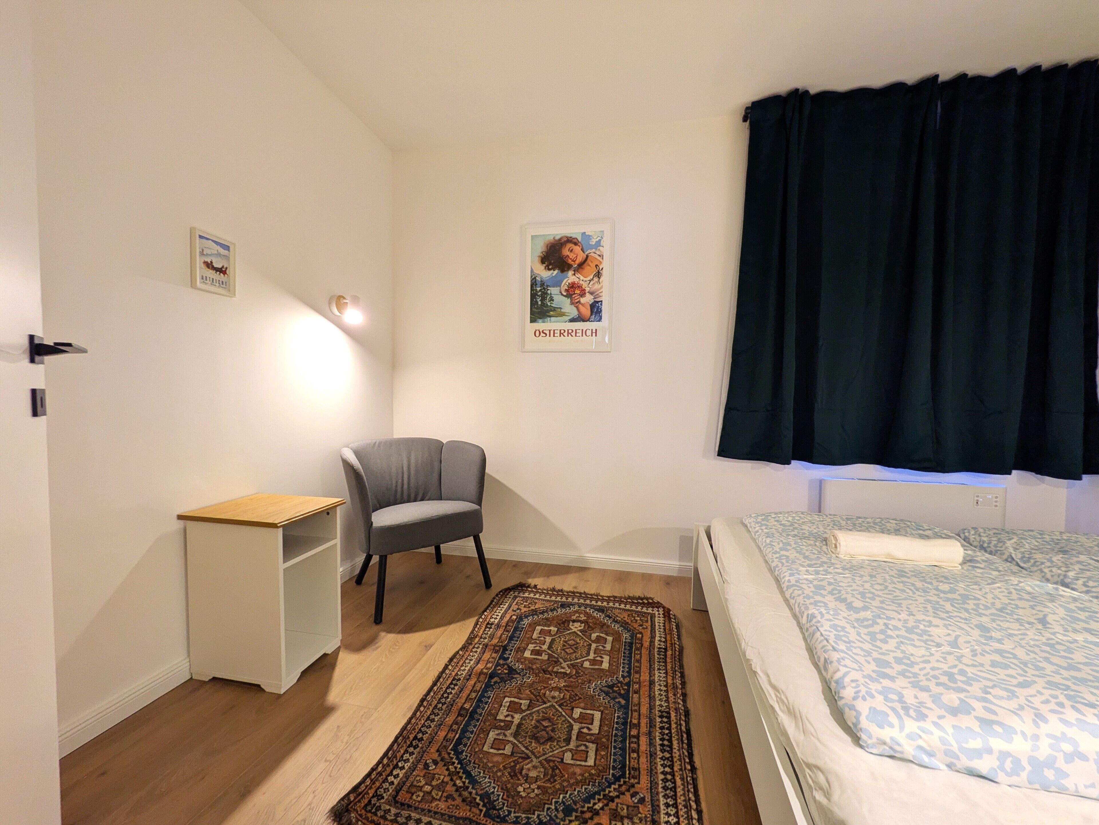 Um quarto pequeno e bem organizado, com uma cama, uma escrivaninha, uma cadeira, um tapete e um quadro na parede.