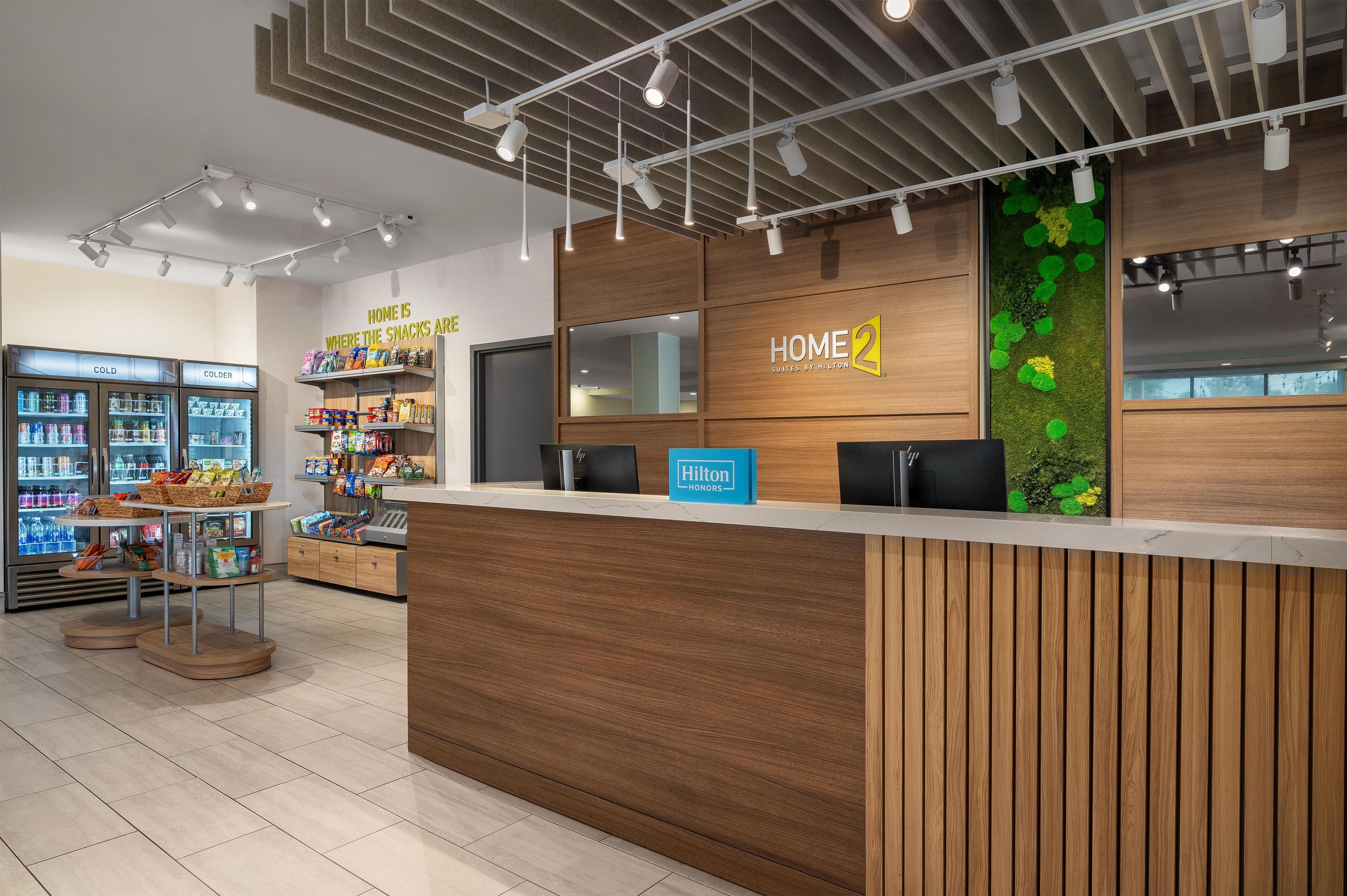 Uma área de recepção de hotel com um balcão de madeira, uma tela digital e uma placa na parede com a inscrição 'HOME 2 Suites by Hilton'.