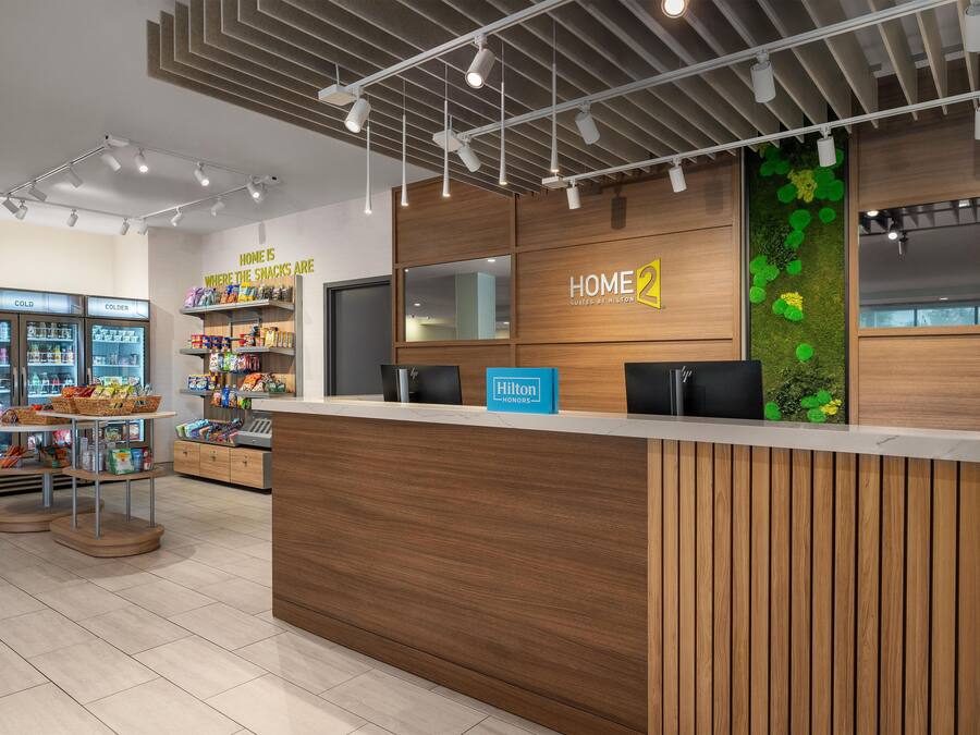 Uma área de recepção de hotel com um balcão de madeira, uma tela digital e uma placa na parede com a inscrição 'HOME 2 Suites by Hilton'.