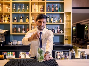 Um bartender preparando uma bebida num bar, com prateleiras cheias de garrafas de bebidas alcoólicas ao fundo.