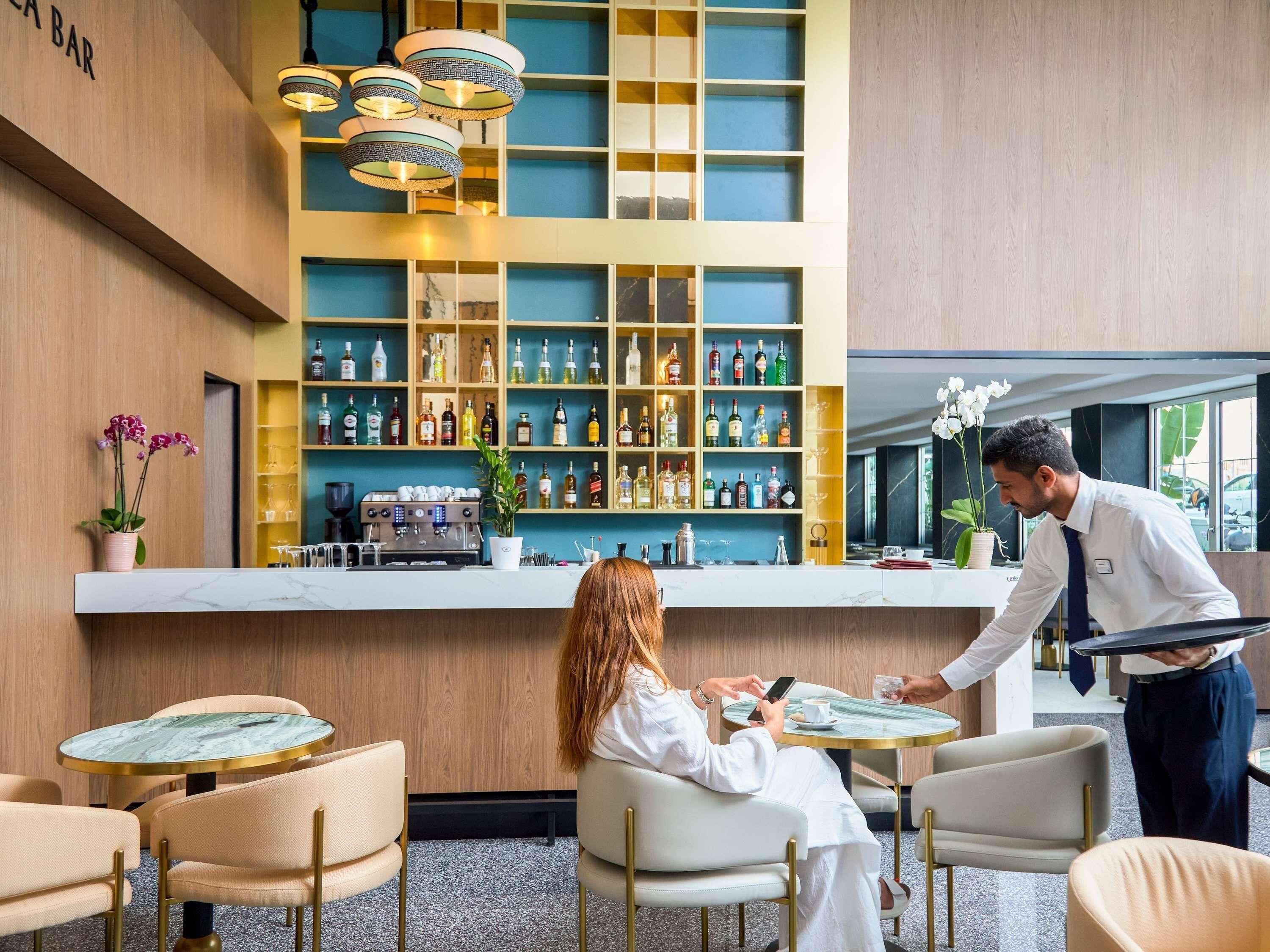 Uma área de bar com balcão, prateleiras com garrafas de bebidas, uma máquina de café e um garçom atendendo um cliente.