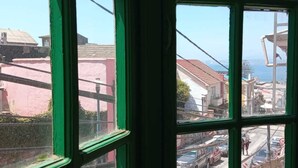 Visão pela janela com molduras verdes, mostrando uma rua com carros estacionados e edifícios ao fundo.