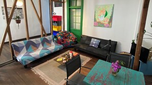 Uma sala de estar aconchegante com uma porta verde, um sofá, uma poltrona colorida, uma pequena mesa com um vaso e um claraboia acima.