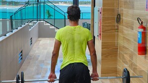 Uma pessoa levantando uma barra em um saguão moderno de hotel, com vista para uma praia e uma escada que leva a uma varanda.