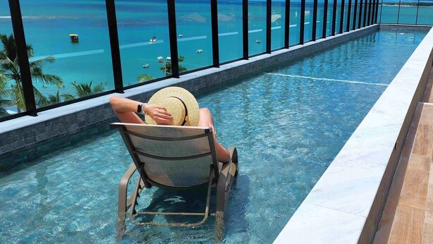 Uma pessoa relaxando em uma piscina com vista para o oceano e palmeiras.