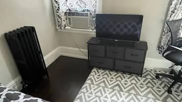 Quarto com radiador, televisão sobre uma cômoda, tapete estampado e uma cadeira.