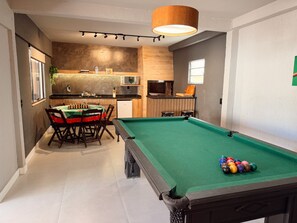 Mesa de bilhar com bolas, área de refeições com mesa e cadeiras, e cozinha com bancada e eletrodomésticos.