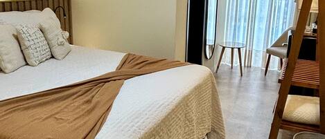 Quarto com uma cama grande, uma cadeira de madeira, um tapete redondo tecido e vista para uma varanda com cortinas.