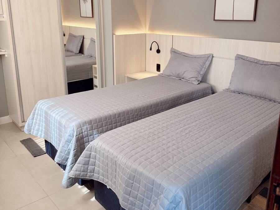 Quarto com duas camas, uma mesinha de cabeceira, uma luminária na parede e uma porta que dá para outro cômodo.