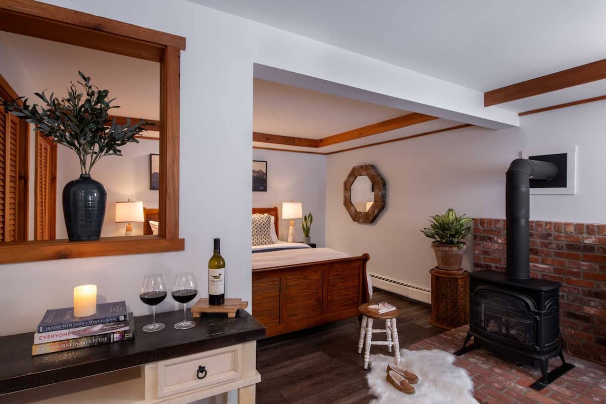 Um quarto aconchegante com uma cama, um banco de madeira, lareira e uma pequena mesa com copos de vinho e uma vela.