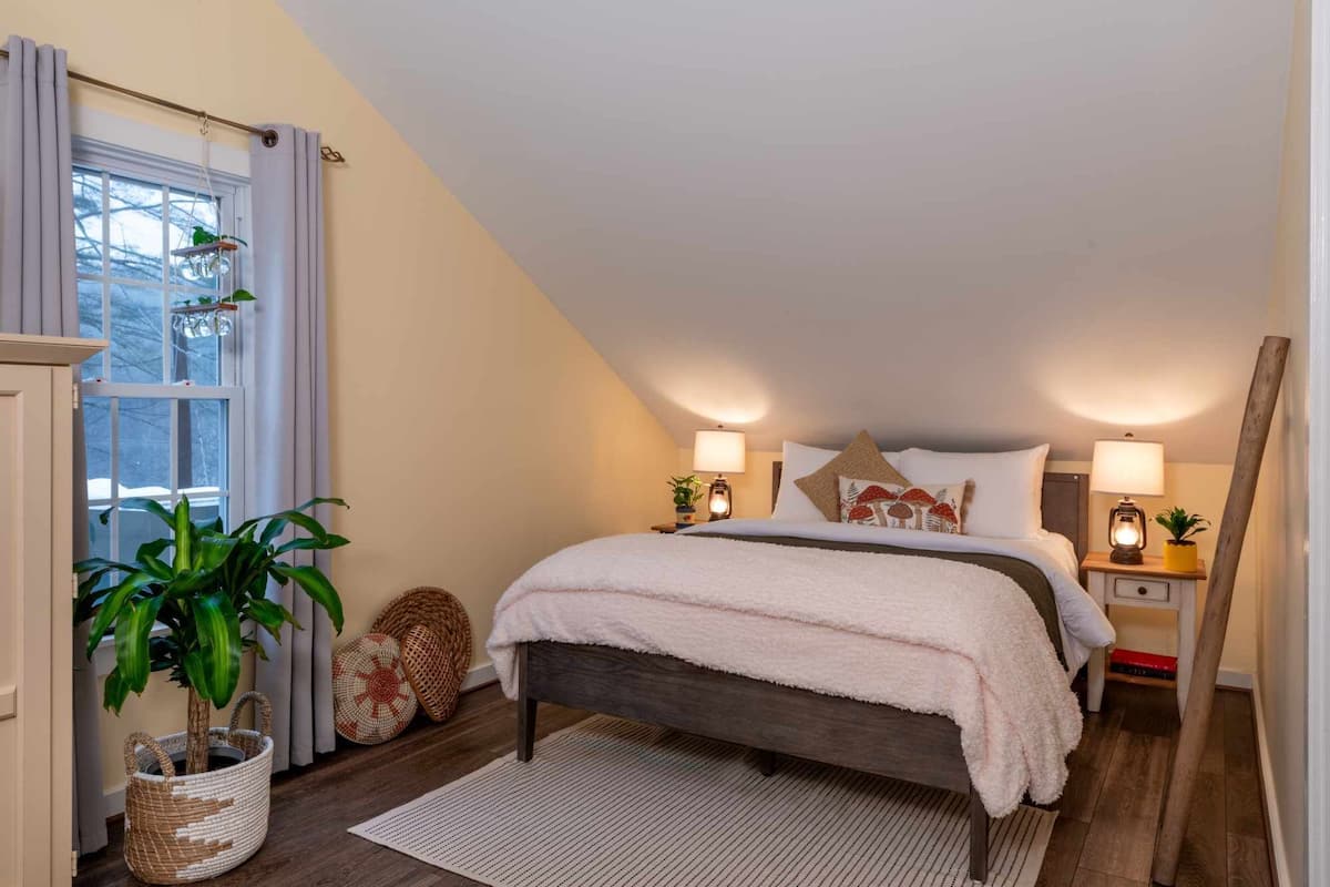 Um quarto com teto inclinado, uma cama com roupa de cama branca, duas luminárias de cabeceira e uma janela com cortinas.