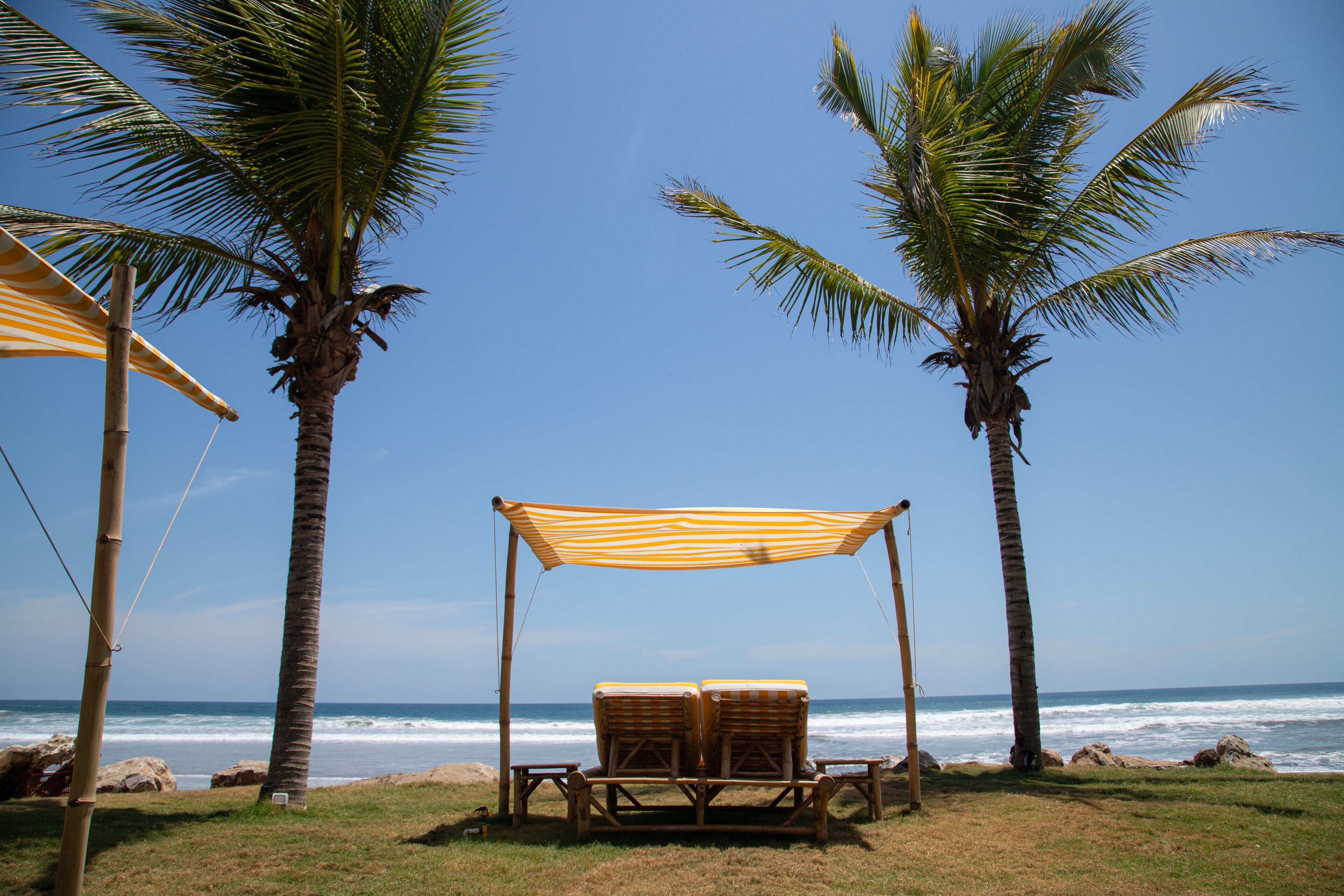 Uma praia com duas espreguiçadeiras sob um toldo listrado, palmeiras e o oceano.