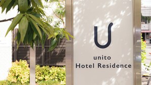 Placa para 'unito Hotel Residence' com um grande logotipo 'U', rodeada por vegetação e um edifício ao fundo.