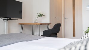 Quarto de hotel moderno com uma cama, televisão fixada na parede, uma pequena mesa de madeira com um vaso de flores e um guarda-roupa.