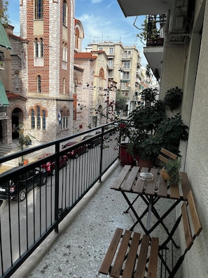 Varanda com mesa e cadeiras de madeira, plantas em vasos e vista para uma rua com carros e edifícios.