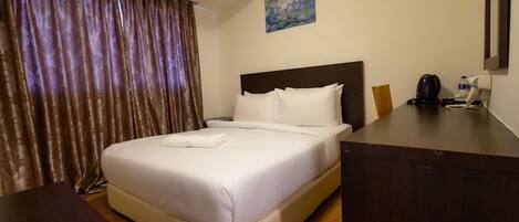 Quarto de hotel com uma cama, uma escrivaninha, uma cadeira, um quadro na parede e uma cortina.