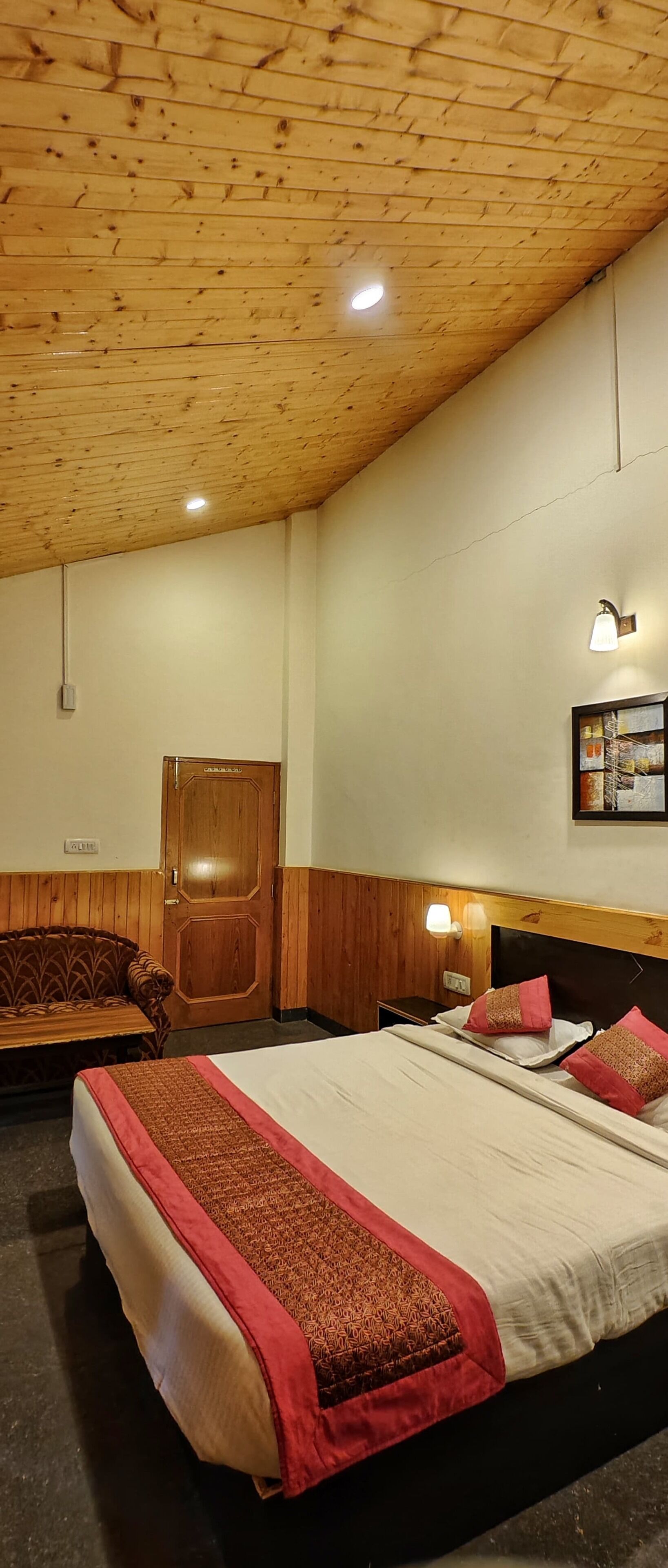 Quarto de hotel com teto de madeira, cama com colcha vermelha e branca, um banco de madeira e luminária fixada na parede.