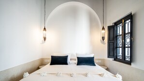 Quarto com cama grande, duas luminárias de cabeceira, janela com grades de ferro decorativas e parede branca.