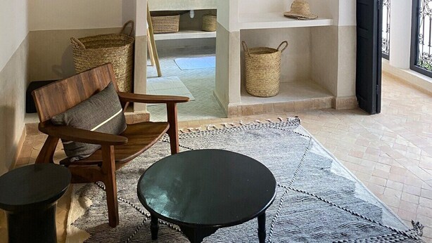 Quarto com uma cadeira de madeira, uma pequena mesa redonda, prateleiras com cestos, um espelho e uma porta em arco.