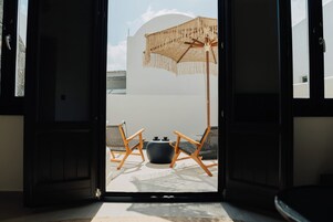 Uma porta aberta dá para uma varanda com um guarda-sol de palha, duas cadeiras de madeira e uma mesa redonda.