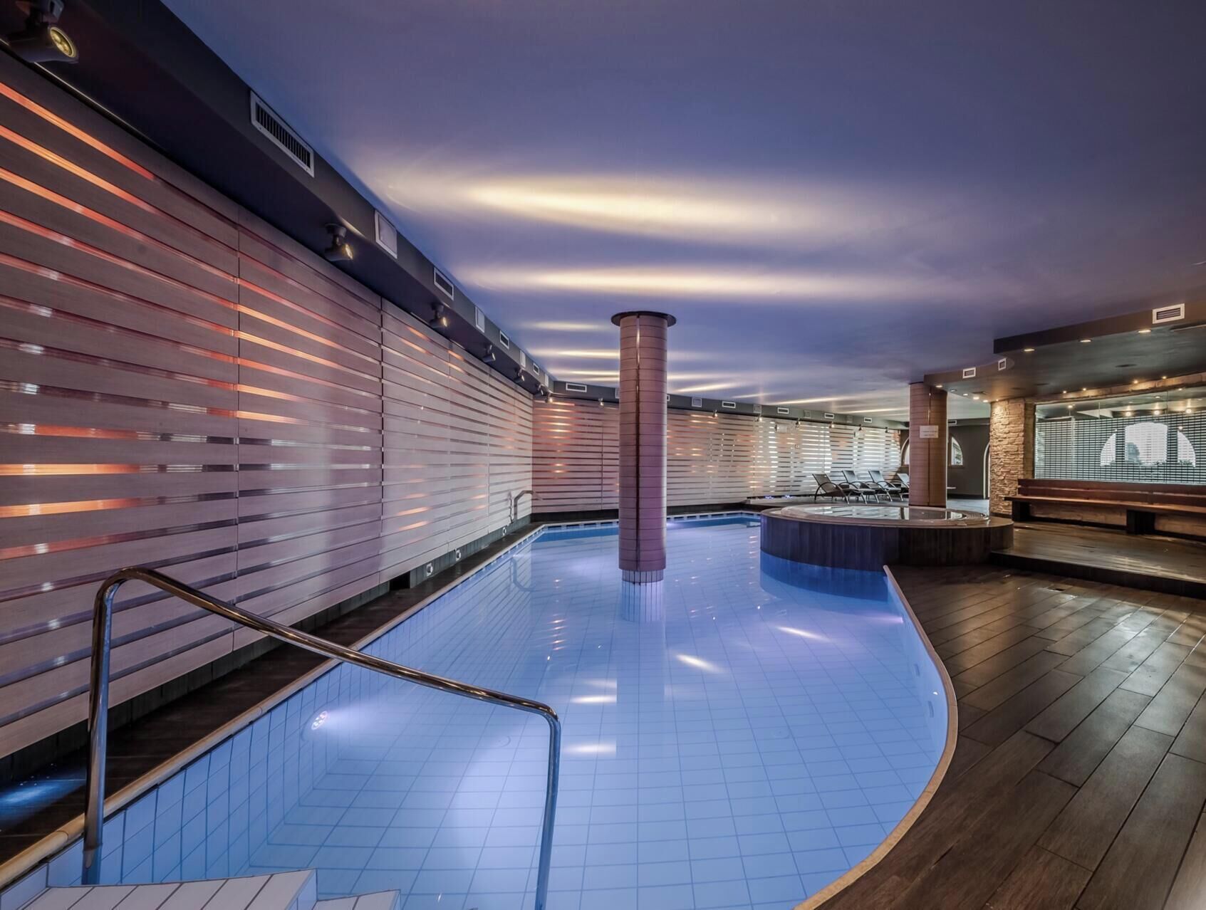 Piscina interior moderna com design curvo, piso de madeira e parede de pedra de um lado.