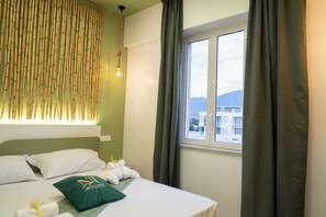 Quarto de hotel com cama, parede verde, janela com cortinas e vista para edifícios.