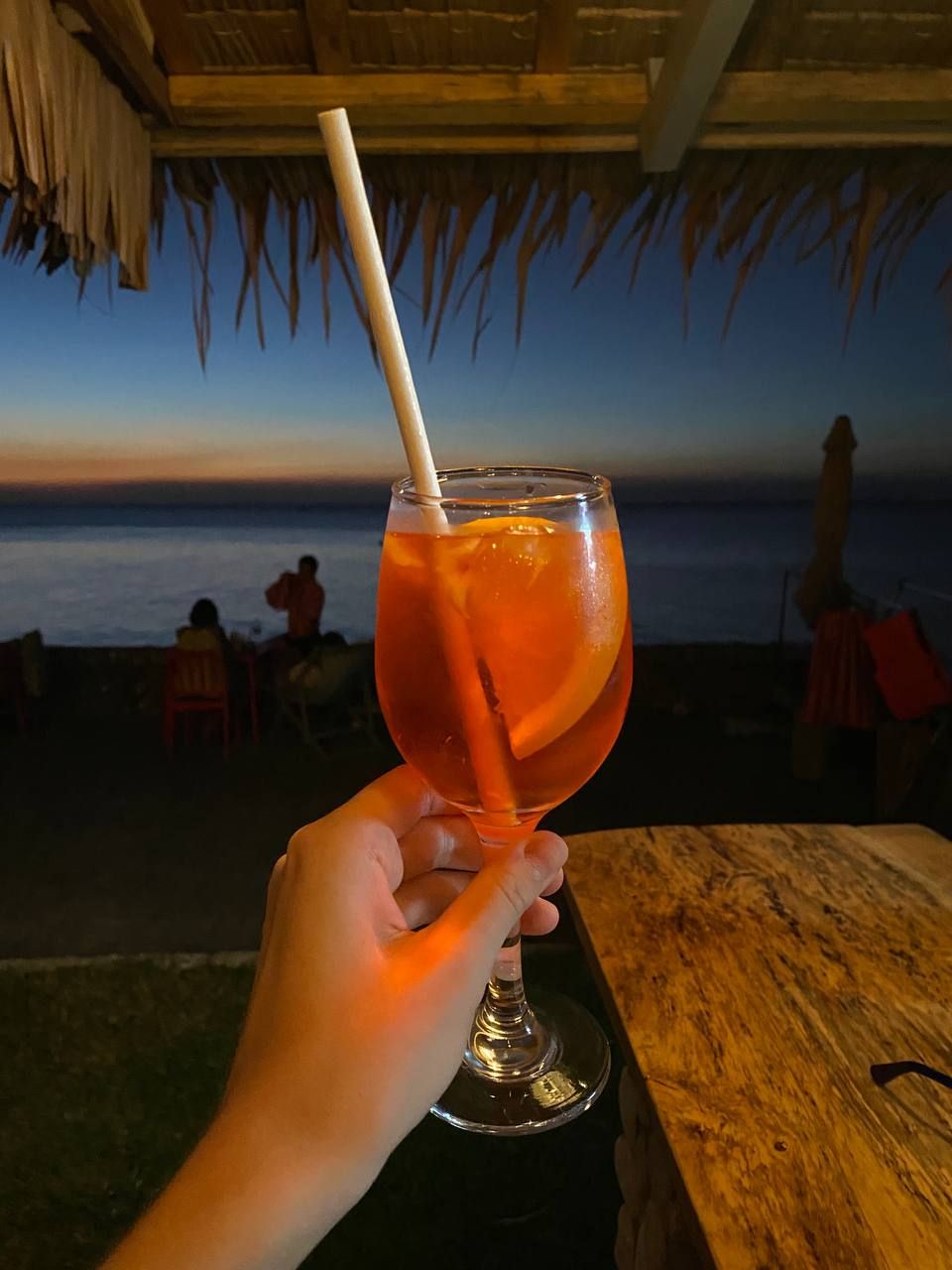 Uma mão segurando um copo com canudo, apoiado em uma mesa de madeira com vista para o oceano ao pôr do sol.