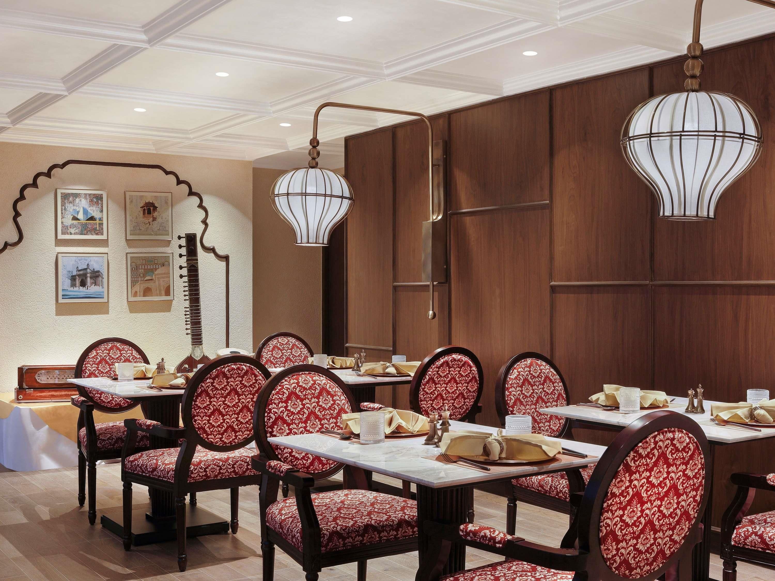 Uma sala de jantar com painéis de madeira, cadeiras estampadas em vermelho, toalhas de mesa brancas e luminárias pendentes.