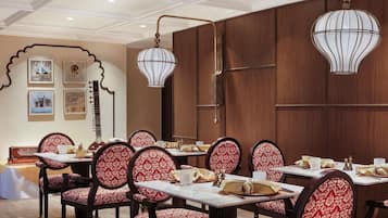 Uma sala de jantar com painéis de madeira, cadeiras estampadas em vermelho, toalhas de mesa brancas e luminárias pendentes.