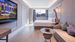 Quarto de hotel moderno com uma cama grande, um sofá, uma mesinha e uma televisão exibindo uma vista urbana.