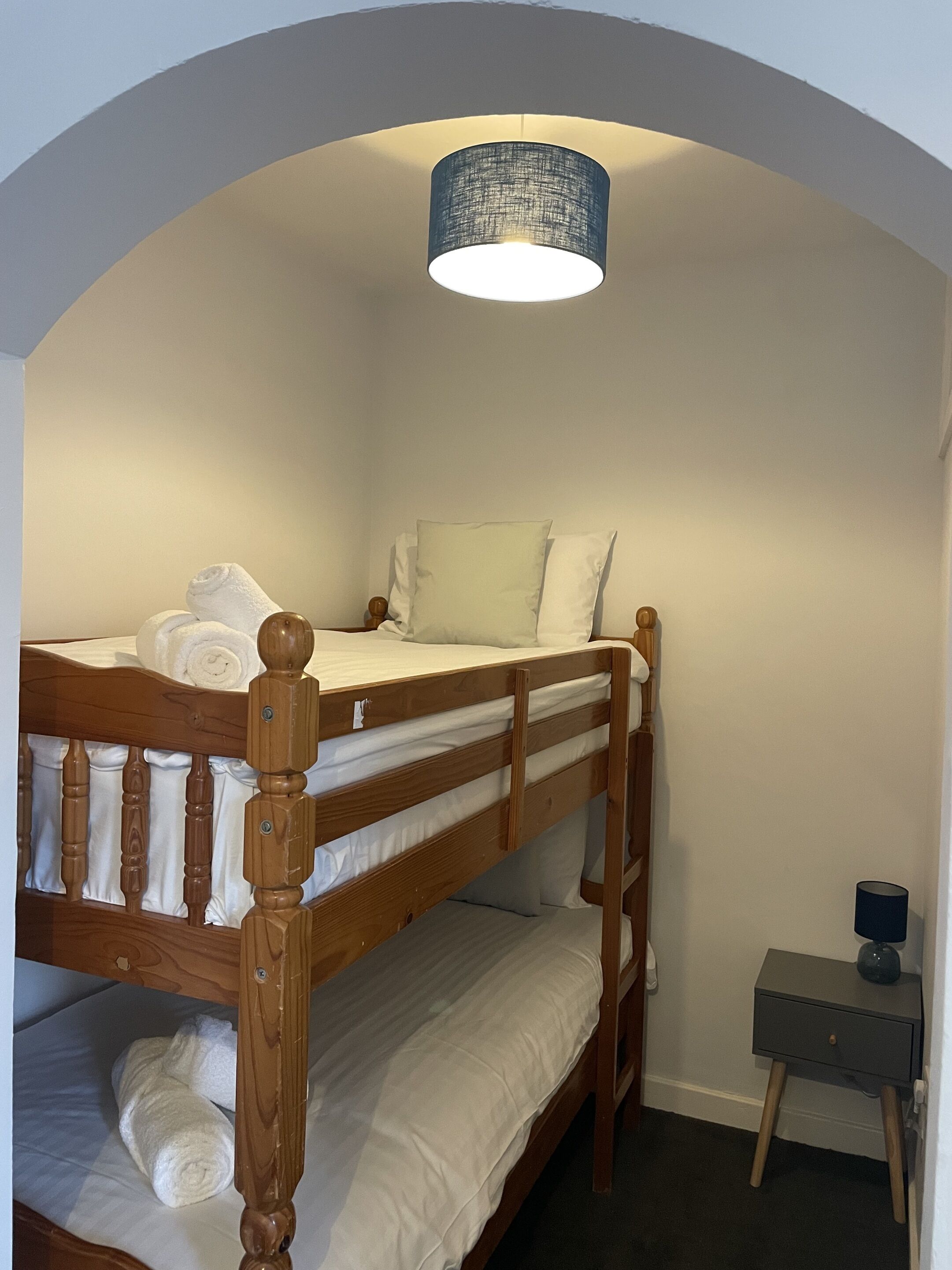 Quarto com beliche, uma luminária sobre a cama, uma mesinha pequena e um arco na parede.