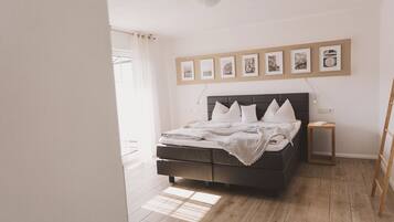Quarto com cama, mesa de cabeceira, piso de madeira e janela com cortinas.