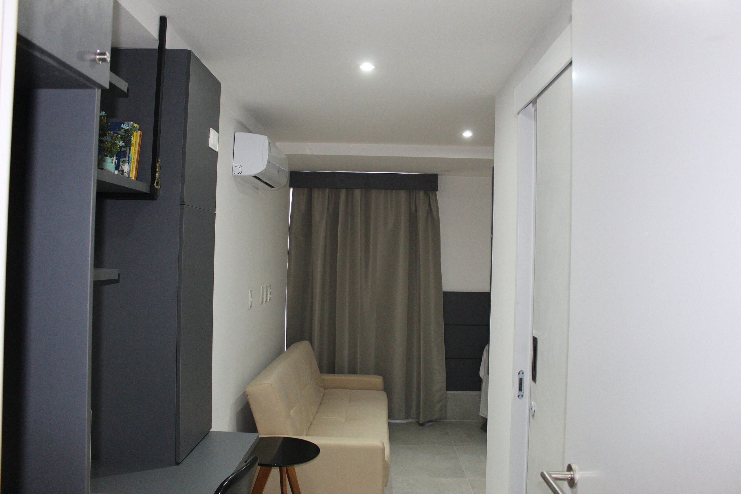 Quarto pequeno com sofá, uma mesinha, guarda-roupa e porta que dá para outro cômodo.