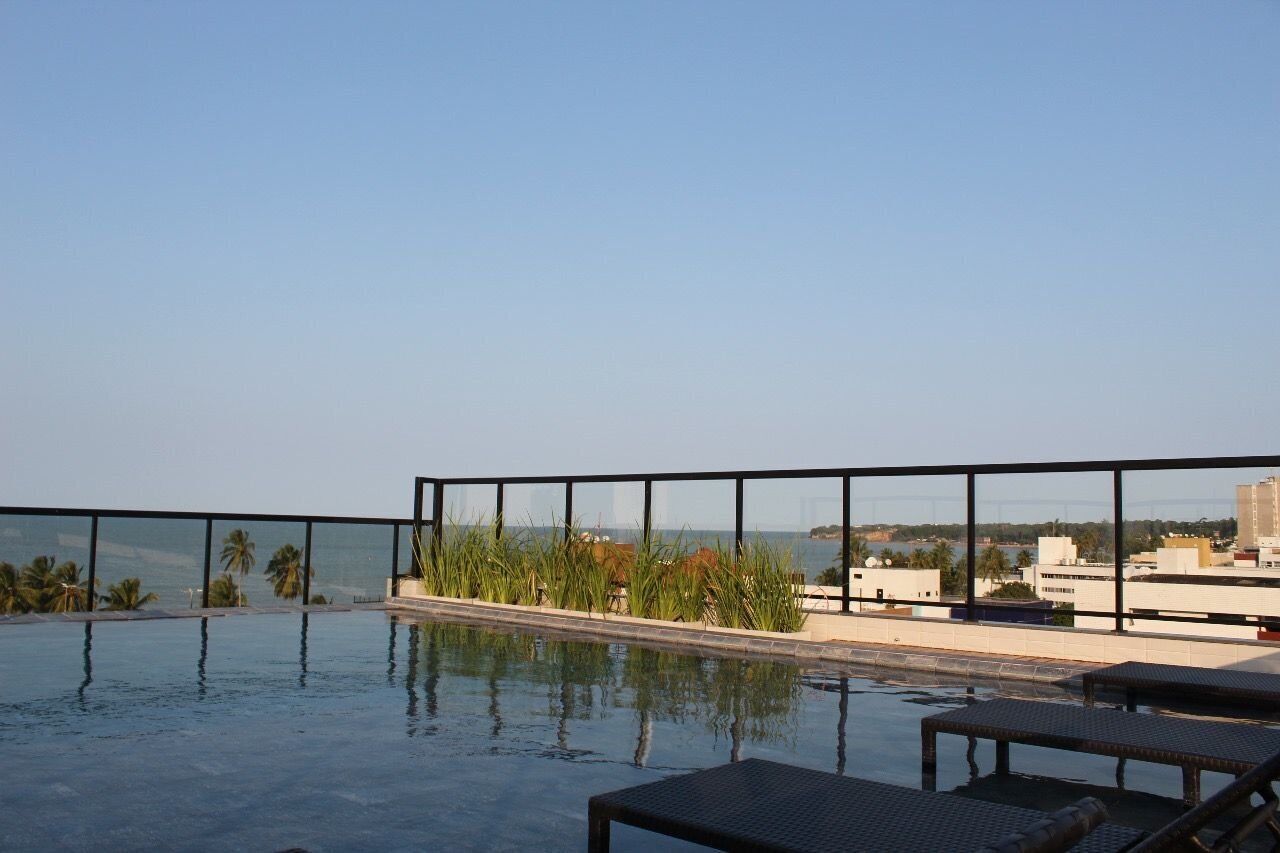Piscina infinita com vista para o oceano e palmeiras, rodeada por espreguiçadeiras e um guarda-corpo de vidro.