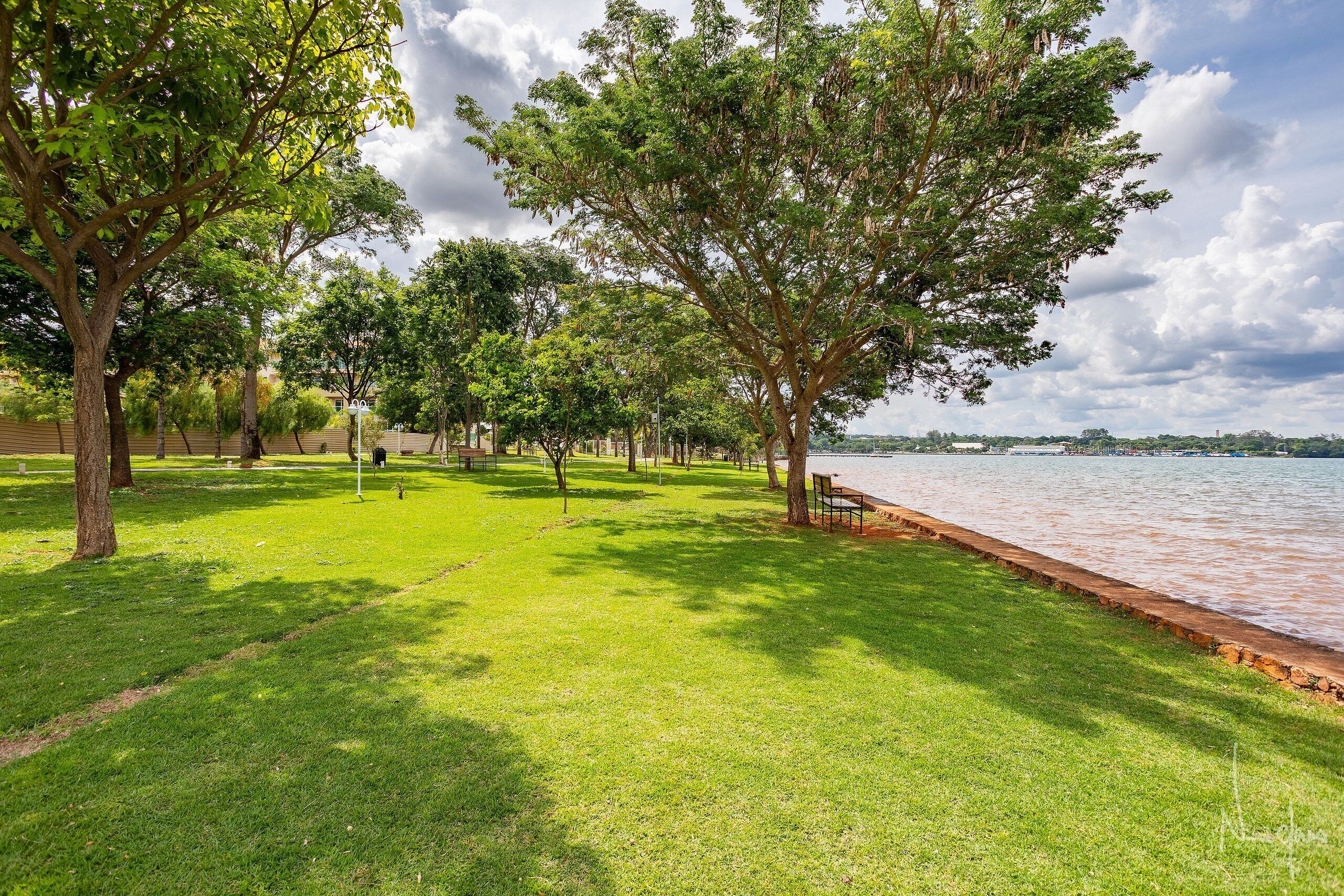 Um parque com gramado, árvores, bancos e um corpo de água.