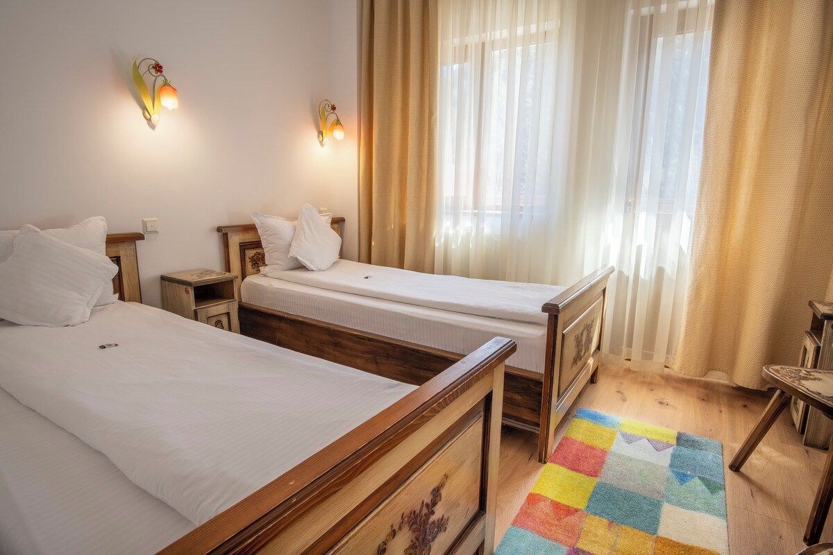 Quarto de hotel com duas camas de solteiro, cabeceiras de madeira, mesinhas de cabeceira e um tapete colorido.