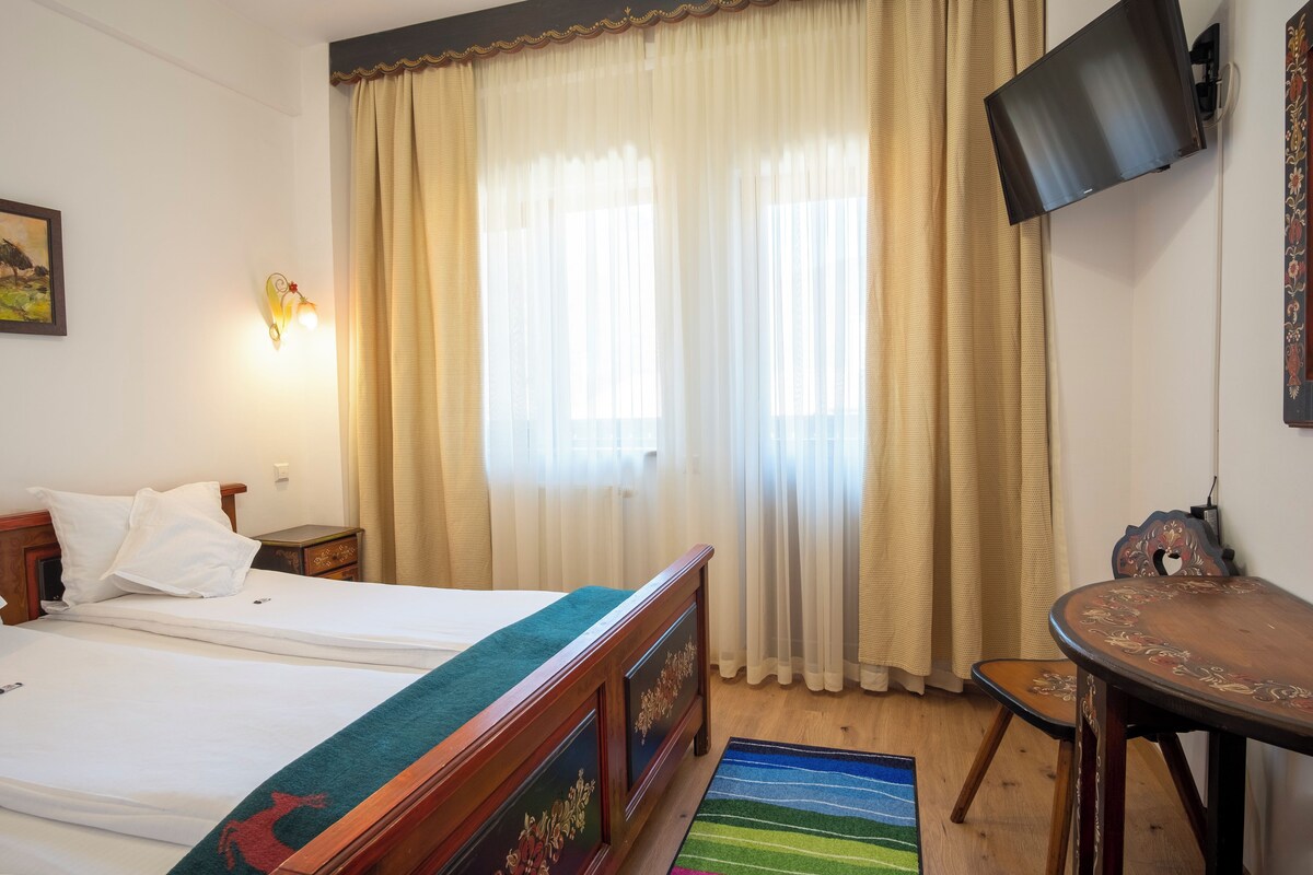 Quarto de hotel com cama, escrivaninha, cadeira, televisão e cortinas.