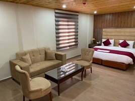 Quarto de hotel com sofá, duas poltronas, mesa de centro de vidro, cama com cabeceira de madeira e janela com persianas.