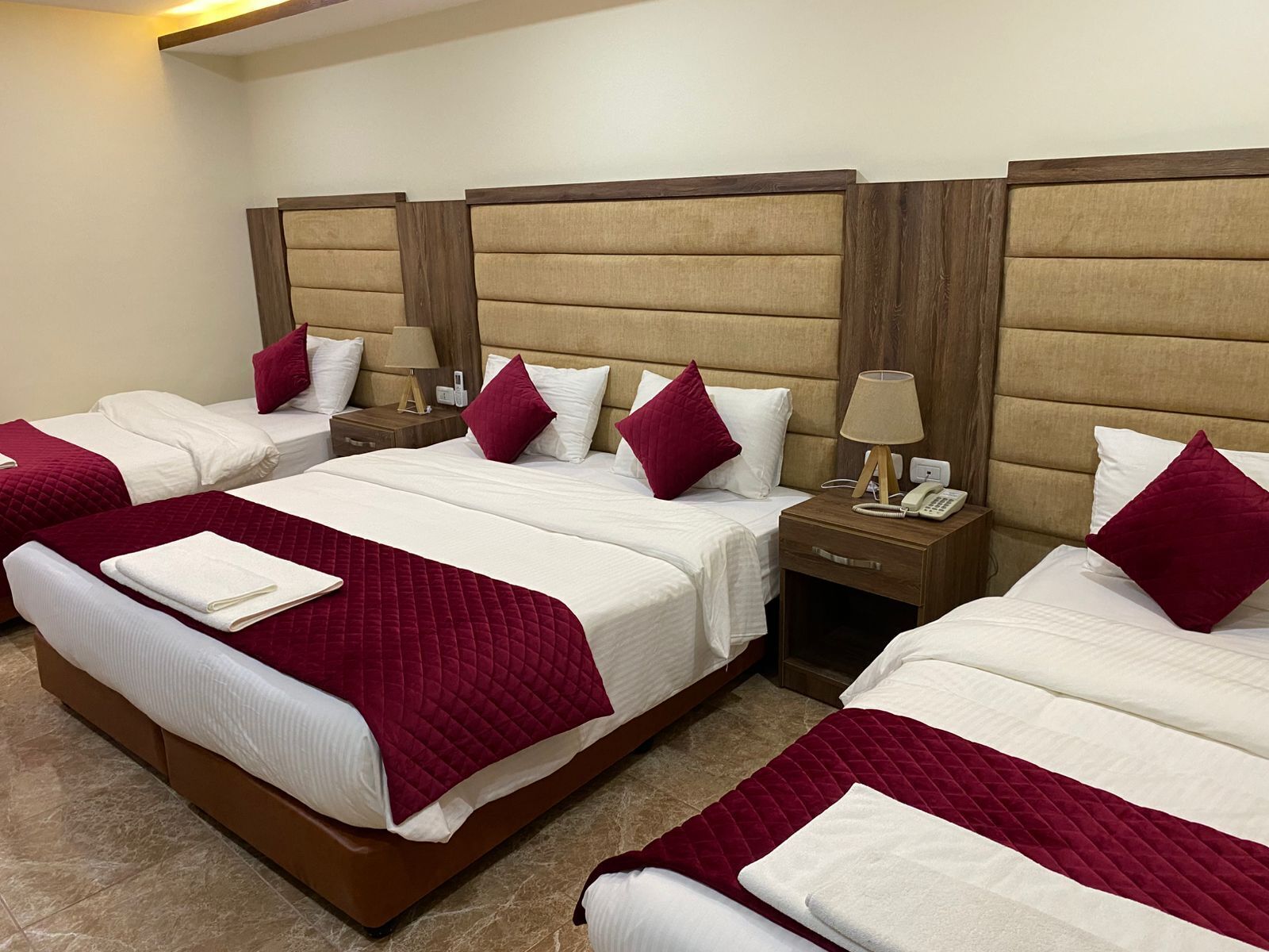 Quarto de hotel com três camas, cada uma com cabeceira de madeira, roupa de cama branca e almofadas vermelhas. Há mesas de cabeceira com abajures e um telefone.